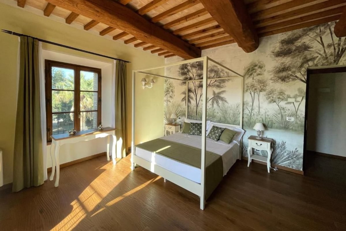 Quarto com cama de dossel, janela com cortinas, mesa de cabeceira e um mural na parede.