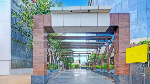 Entrada de um edifício moderno com galeria coberta, plantas em vasos e uma placa amarela.