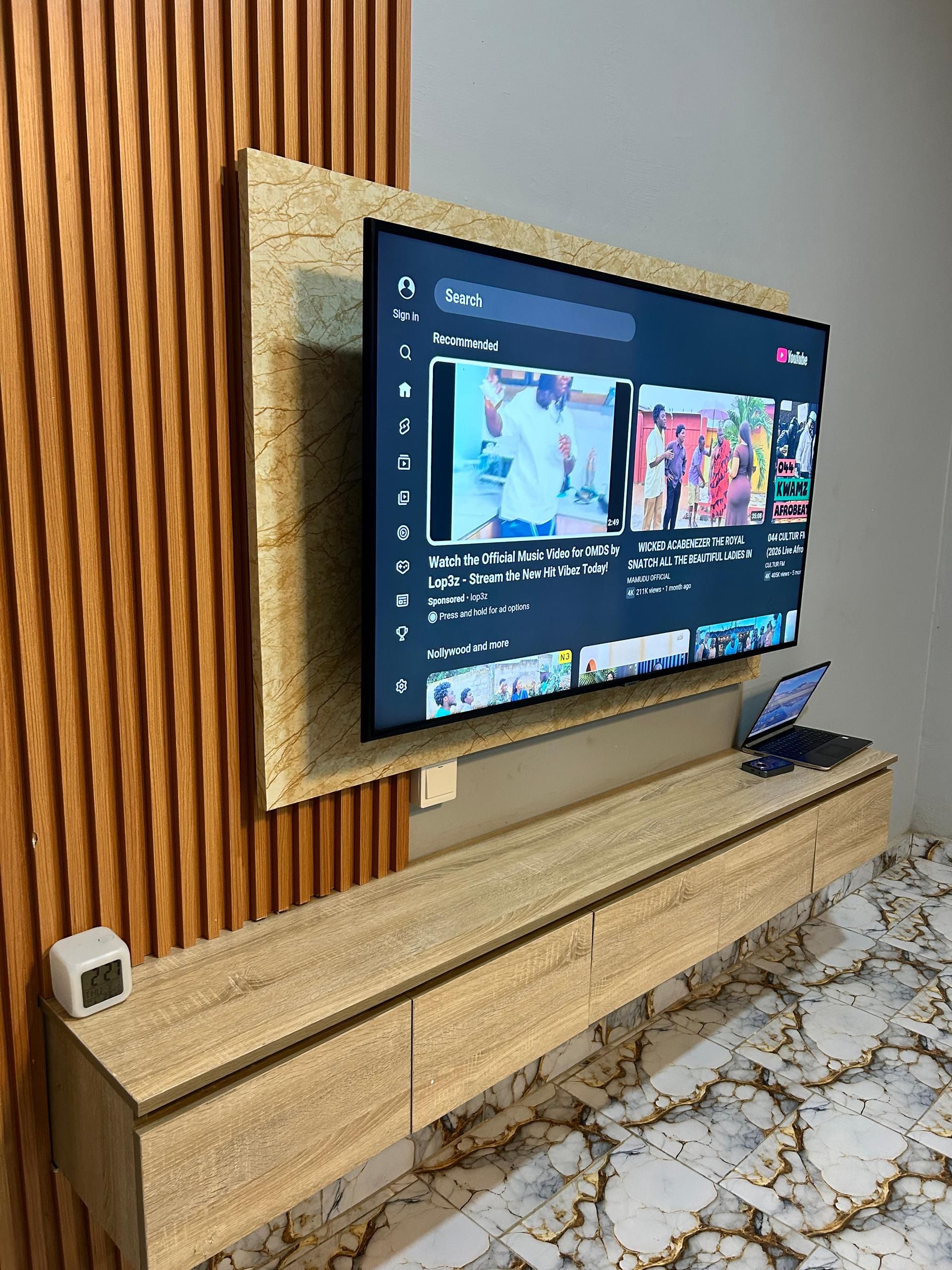 Televisão de tela plana fixada em parede de madeira, exibindo uma página do YouTube com diversas miniaturas de vídeos.
