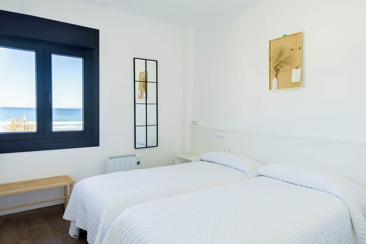 Quarto com duas camas, janela com vista para o mar, mesa de cabeceira e espelho fixado na parede.