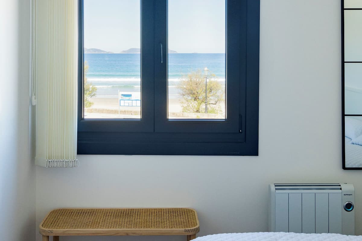 Quarto com uma janela grande, cama, banqueta e radiador.