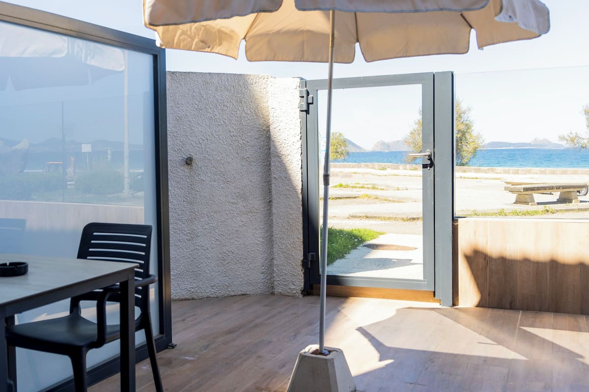 Um pátio aberto com mesa e cadeiras, um guarda-sol bege e vista para o mar.