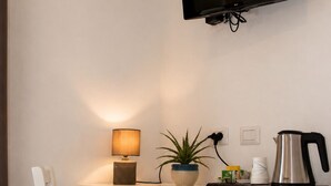 Quarto pequeno com escrivaninha branca, cadeira, televisão na parede, ar condicionado, luminária, planta, chaleira e mini-geladeira com bebidas.