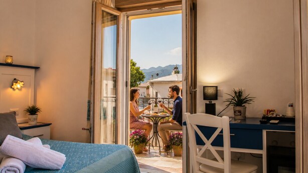 Quarto com cama, mesa onde duas pessoas jantam, cadeira, escrivaninha e vista para o exterior.