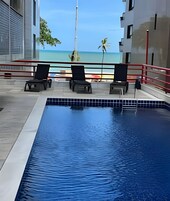 Uma piscina com espreguiçadeiras, vista para a praia e uma construção alta ao fundo.