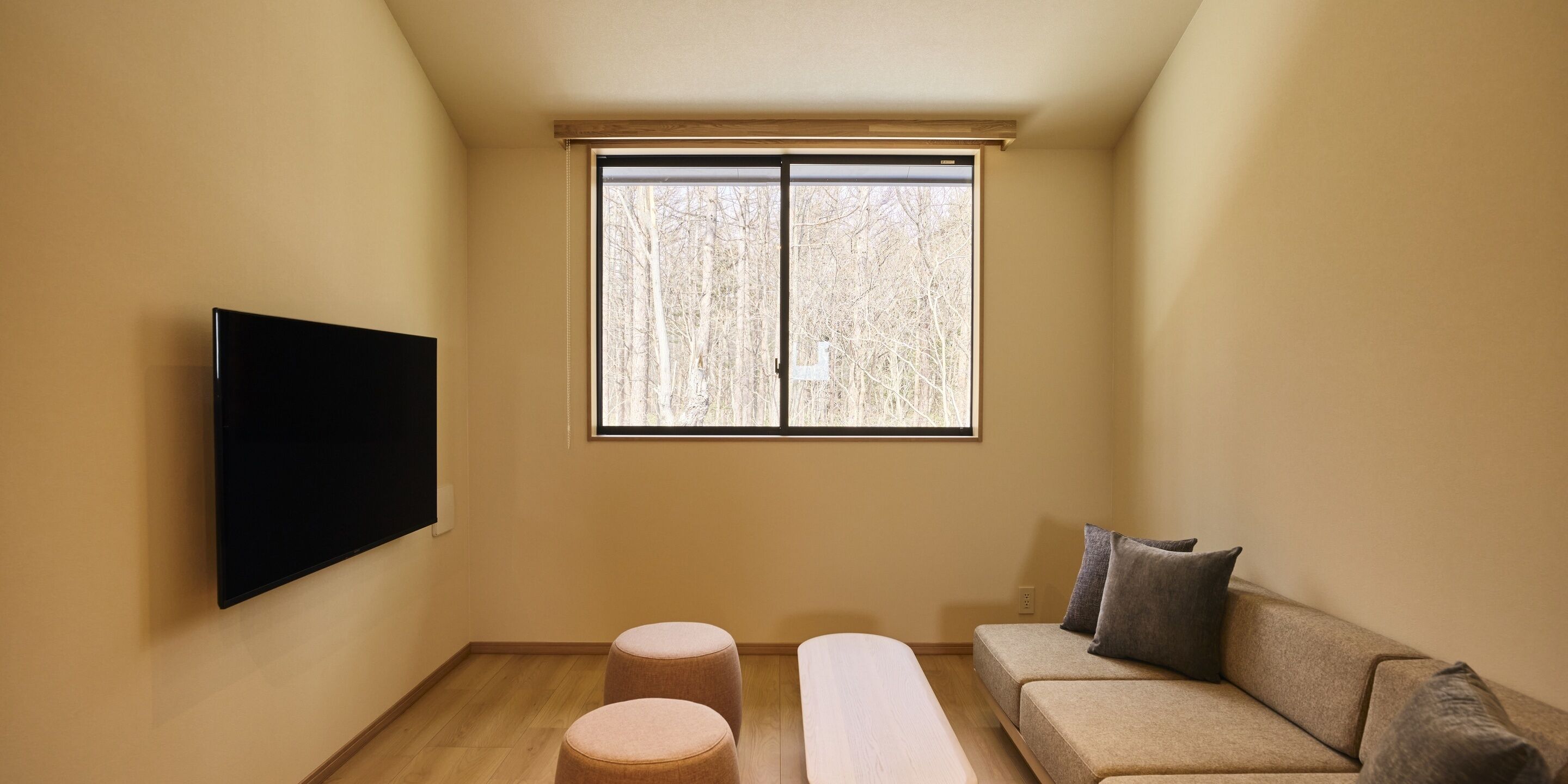 Um quarto com televisão de tela plana na parede, uma janela com vista para árvores, um sofá bege com almofadas e dois pufes redondos.