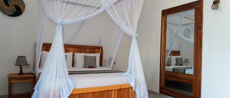 Quarto com cama de madeira, roupa de cama branca, mesa de cabeceira com abajur, espelho grande e um pendurar de parede em tecido.