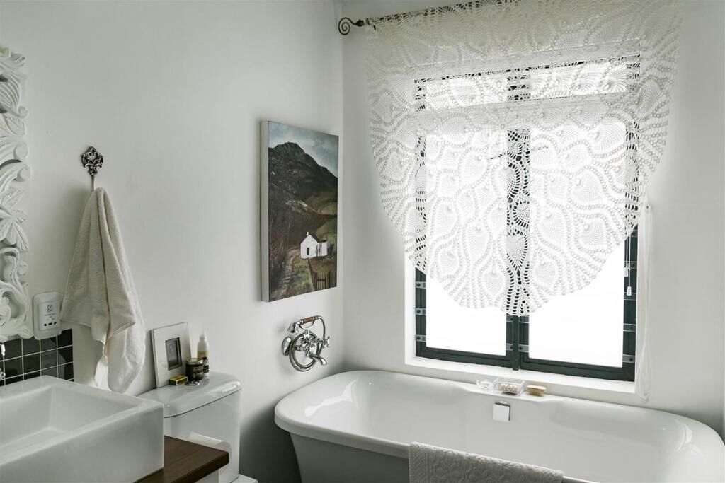 Um banheiro com uma banheira branca, uma toalha pendurada na parede, um quadro de uma montanha e uma janela com cortinas de renda.