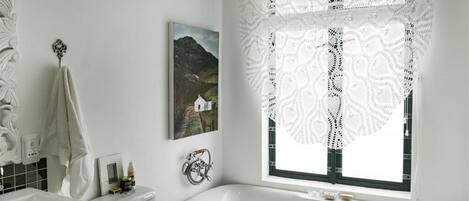 Um banheiro com uma banheira branca, uma toalha pendurada na parede, um quadro de uma montanha e uma janela com cortinas de renda.