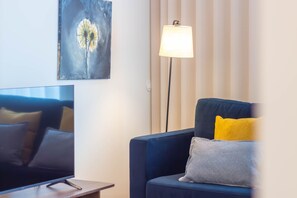 Uma sala de estar moderna com televisão de tela plana, um sofá azul escuro com almofada estampada, um abajur e uma obra de arte abstrata na parede.