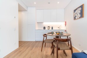 Uma cozinha compacta com armários brancos, uma mesa de jantar com cadeiras e um quadro na parede.