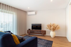 Uma sala de estar moderna com televisão de tela plana, mesa consola de madeira, uma janela grande com cortinas translúcidas e um vaso com plantas secas.