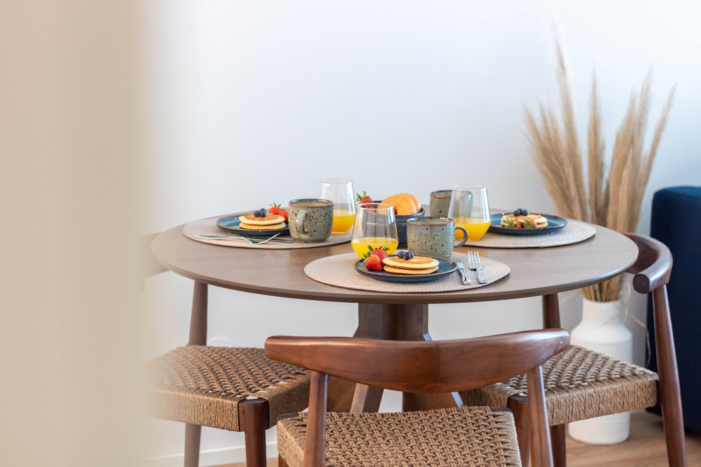 Uma mesa de jantar redonda com um jogo americano, uma pilha de panquecas, um copo de suco de laranja e uma caneca.