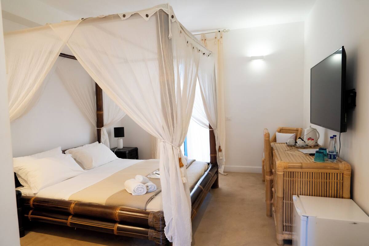 Quarto com cama de dossel, televisão, mesinha com cadeira e mesa de cabeceira com abajur.
