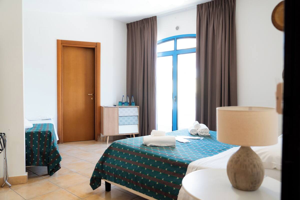 Quarto de hotel com cama, mesa de cabeceira, luminária, janela com cortinas, porta e pequena mesa com toalha.