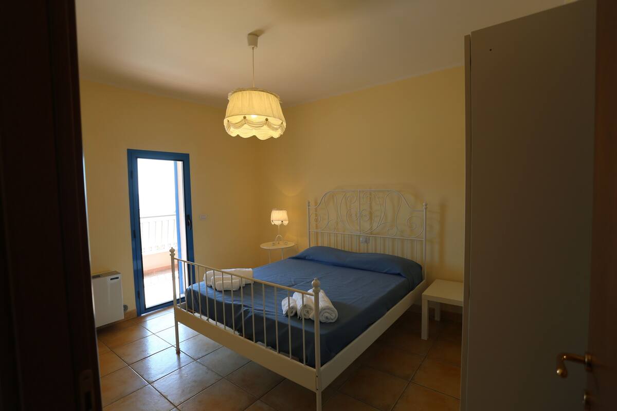 Quarto com cama, mesa de cabeceira, luminária e porta.