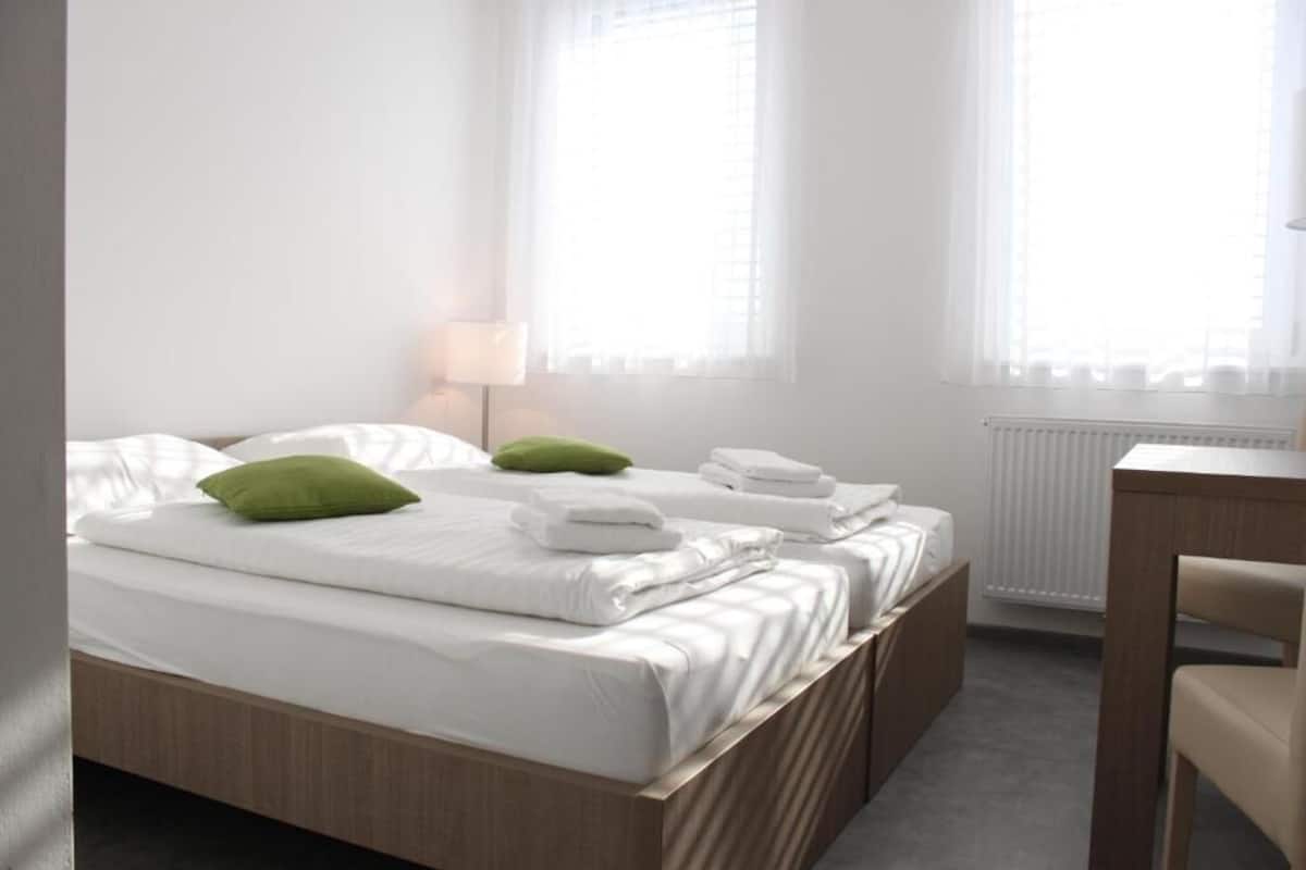 Quarto de hotel com duas camas, uma escrivaninha e um radiador.