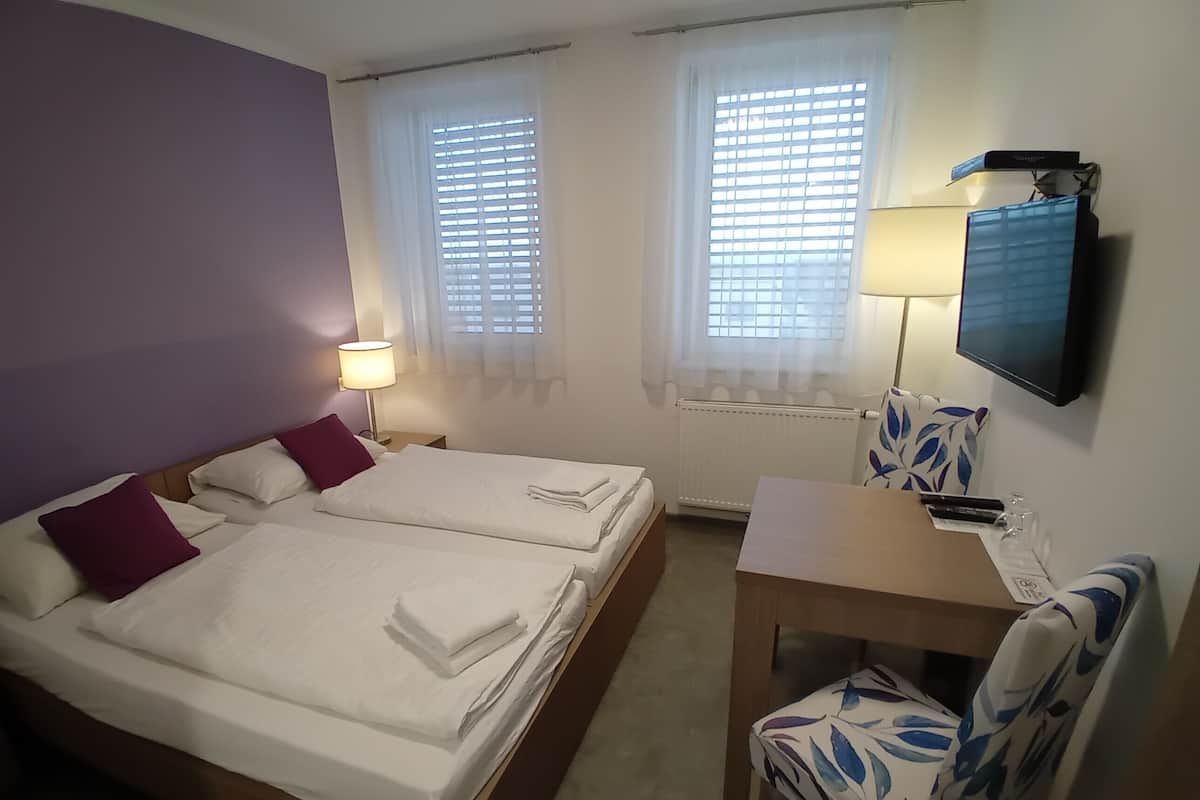 Quarto de hotel com duas camas, uma escrivaninha, uma cadeira, uma televisão e duas janelas com persianas.