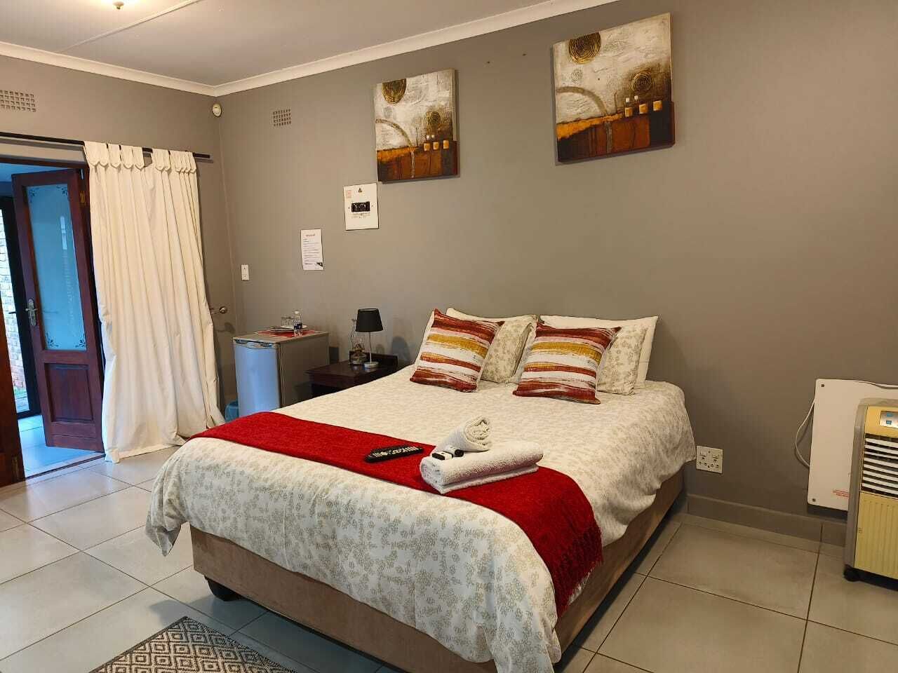 Cama bem arrumada com manta vermelha, um controle remoto e uma toalha sobre ela. Duas pinturas na parede, uma mesa de cabeceira com abajur e um aquecedor no chão.
