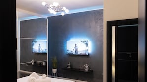 Quarto de hotel moderno com uma cama grande, televisão fixada na parede, uma pequena planta e um espelho.