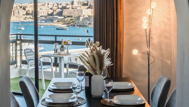Uma sala de jantar com uma mesa posta para dois, um vaso com flores e vista para o mar e edifícios.