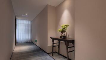 Um corredor com piso de madeira, um vaso com planta sobre uma mesa e uma janela com cortinas leves.