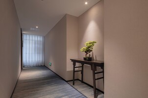 Um corredor com piso de madeira, um vaso com planta sobre uma mesa e uma janela com cortinas leves.