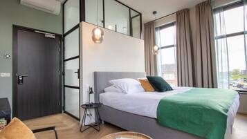 Quarto de hotel com cama, uma pequena mesa com uma revista, escrivaninha, cadeira, janela com cortinas e aparelho de ar condicionado.