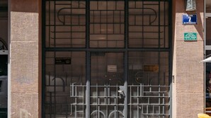 Entrada de um edifício com portão de metal, placas de recepção e uma pequena placa indicando um local próximo.