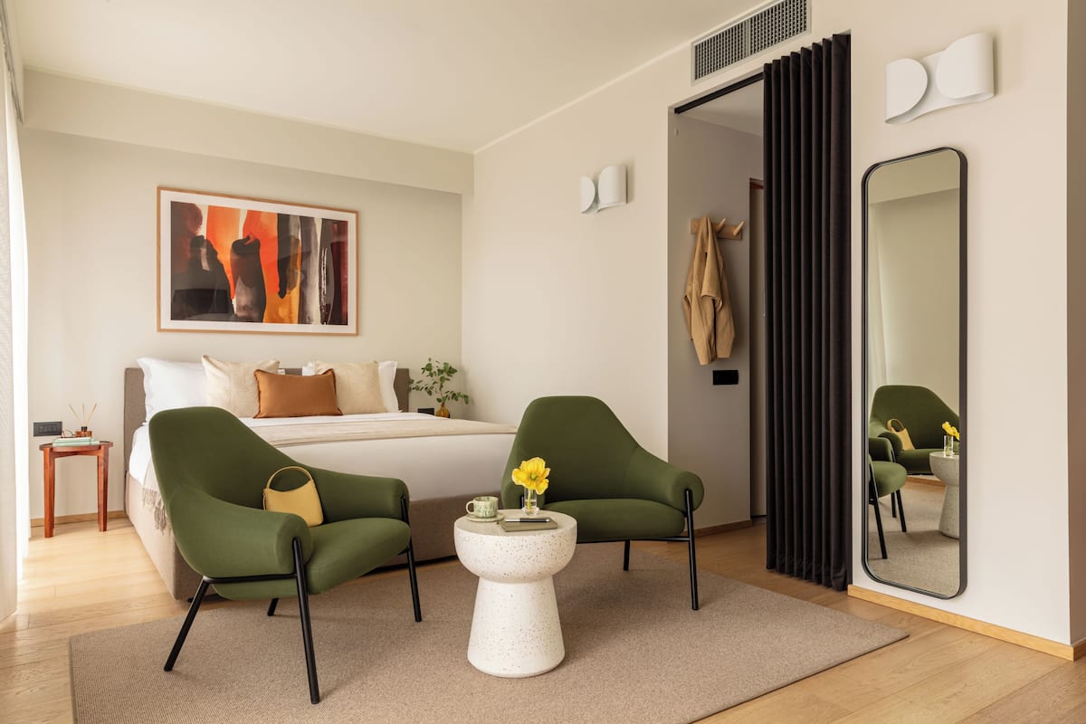 Quarto de hotel moderno com uma cama, duas cadeiras, uma mesinha de apoio, um espelho e um quadro na parede.