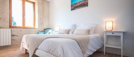 Quarto com uma janela grande, uma cama com roupa de cama branca, uma mesa de cabeceira e um quadro na parede.