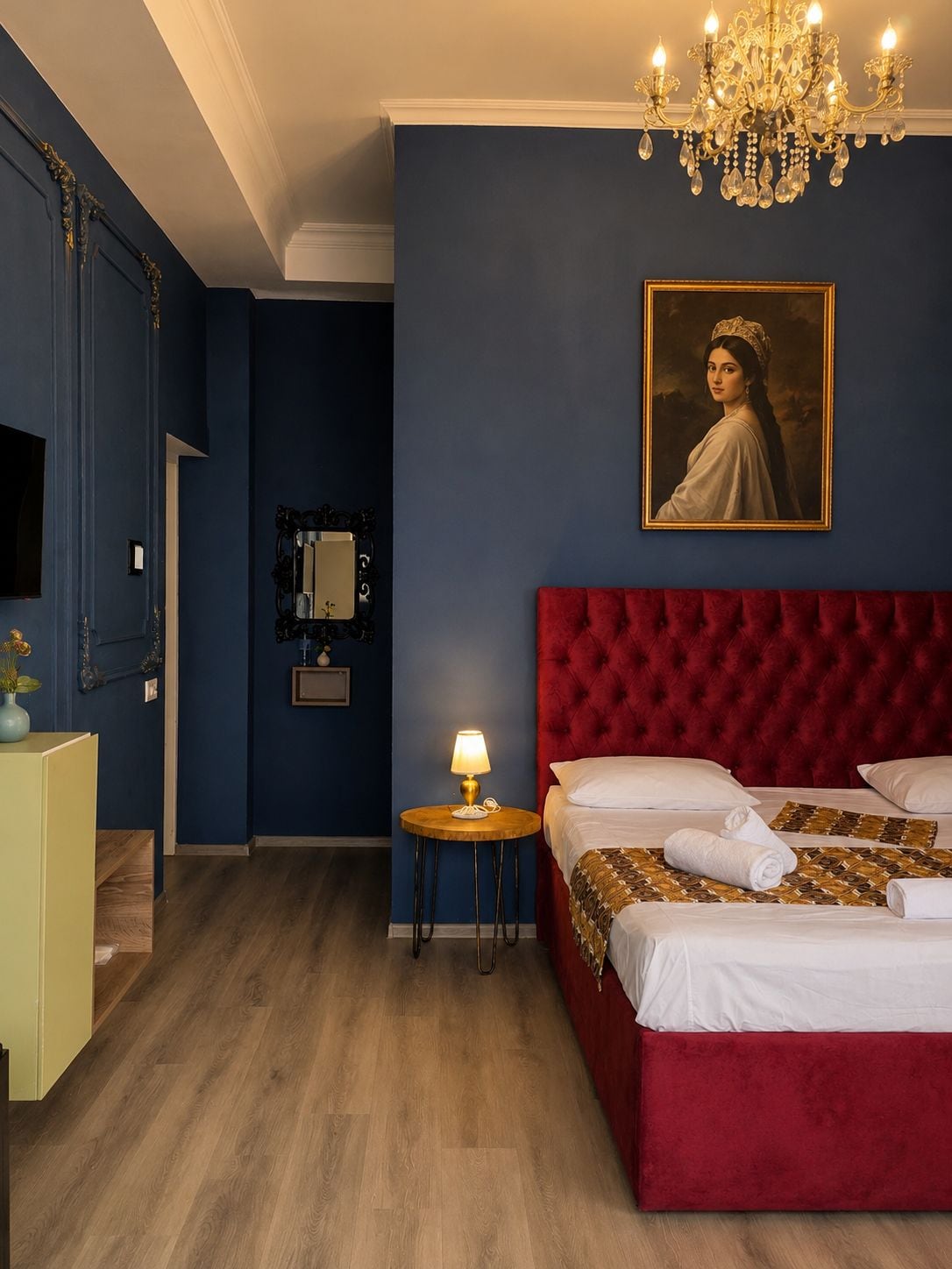 Quarto de hotel com cabeceira estofada em vermelho, cama com roupa de cama branca, mesa lateral redonda com abajur, um quadro na parede e um lustre.