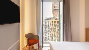 Quarto de hotel com uma cama, uma cadeira, uma televisão presa à parede e uma janela grande com cortinas.