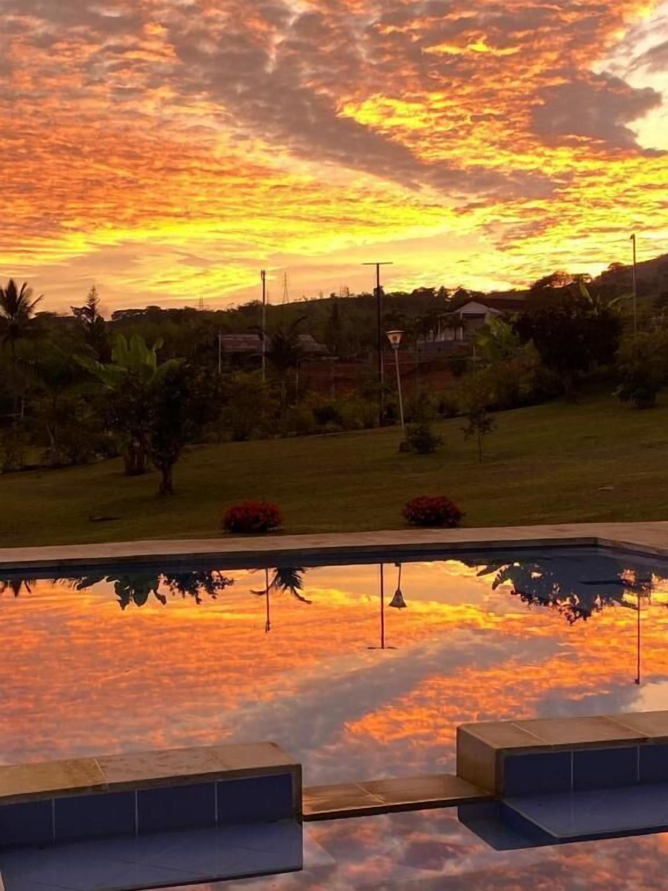 Piscina com vista para o pôr do sol, rodeada por vegetação e alguns edifícios ao fundo.