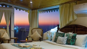 Quarto de hotel com uma cama, duas cadeiras, uma mesa com abajur e vista para o pôr do sol.