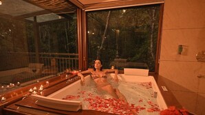 Uma pessoa relaxando em uma banheira cheia de água e pétalas de rosa, com velas e um cesto de flores por perto.