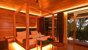 Uma cama de madeira com dossel e vista para um lago, uma mesinha e um sofá num quarto com amplas janelas.