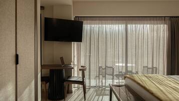 Quarto de hotel com cama, escrivaninha com laptop, cadeira, televisão e vista através de cortinas translúcidas.