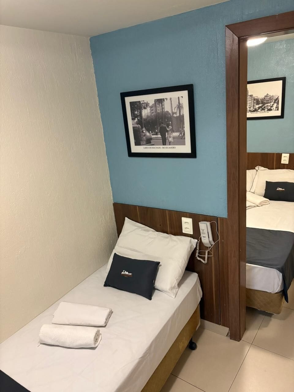 Quarto de hotel com uma cama de solteiro, mesa de cabeceira, telefone, quadro na parede e um espelho refletindo parte do quarto.