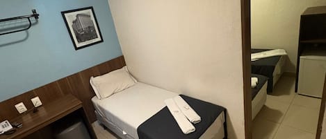 Quarto de hotel compacto com uma cama de solteiro, uma pequena escrivaninha com telefone, um cesto de lixo e um quadro na parede.