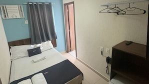Quarto de hotel com cama, pequena escrivaninha, aquecedor, cabideiro e porta que dá para outro cômodo.