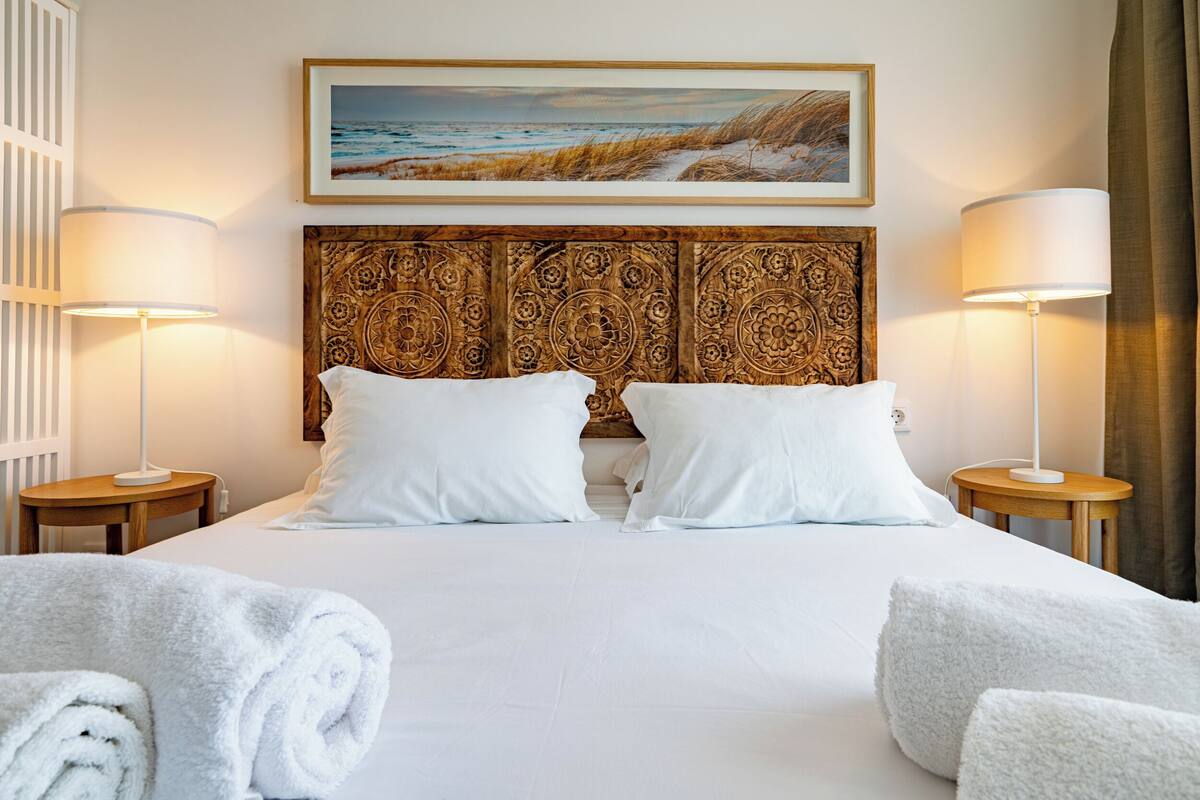 Quarto de hotel com uma cama grande, duas luminárias de cabeceira, um painel de cabeceira decorativo, um quadro pendurado acima da cama e toalhas enroladas sobre a cama.