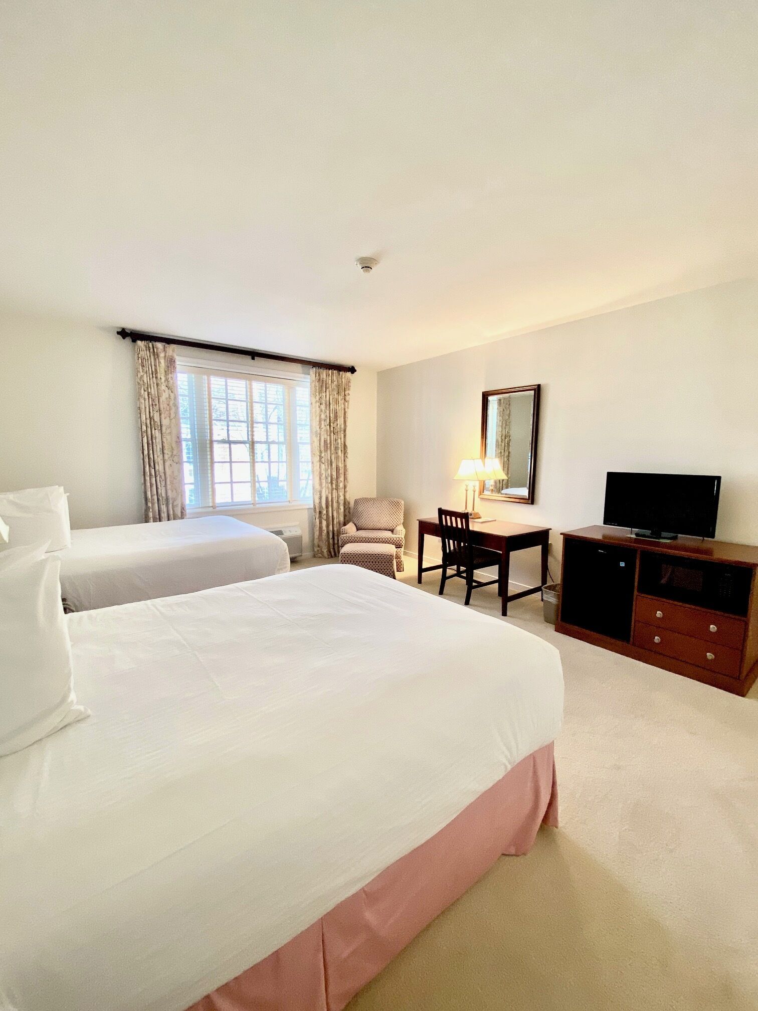Quarto de hotel com duas camas, uma escrivaninha com cadeira, televisão e janela com cortinas.