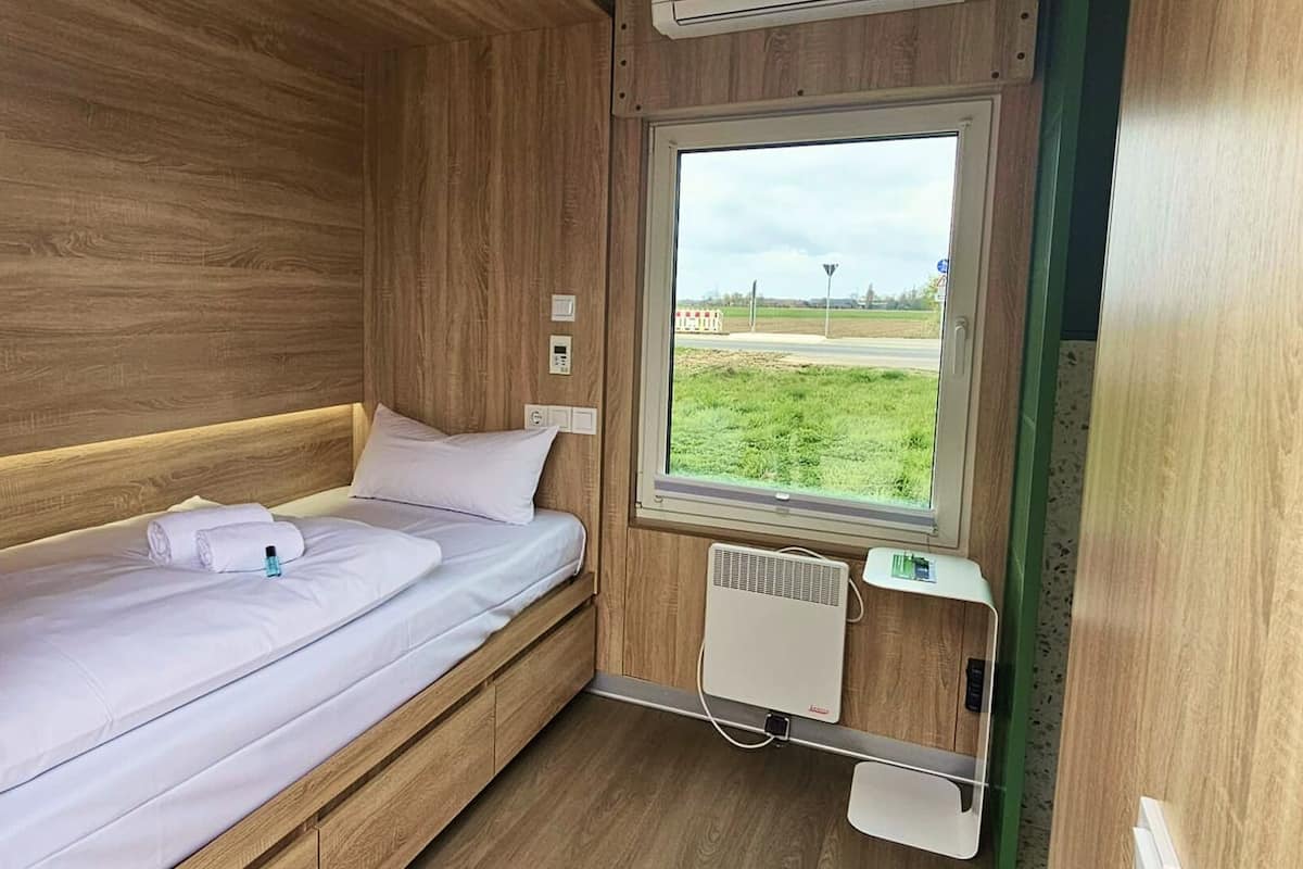 Uma cama de solteiro com cabeceira de madeira, janela com vista para vegetação e ar condicionado fixado na parede.