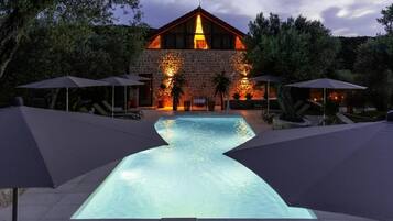 Um edifício de pedra com piscina à frente, cercado por guarda-sóis e árvores.
