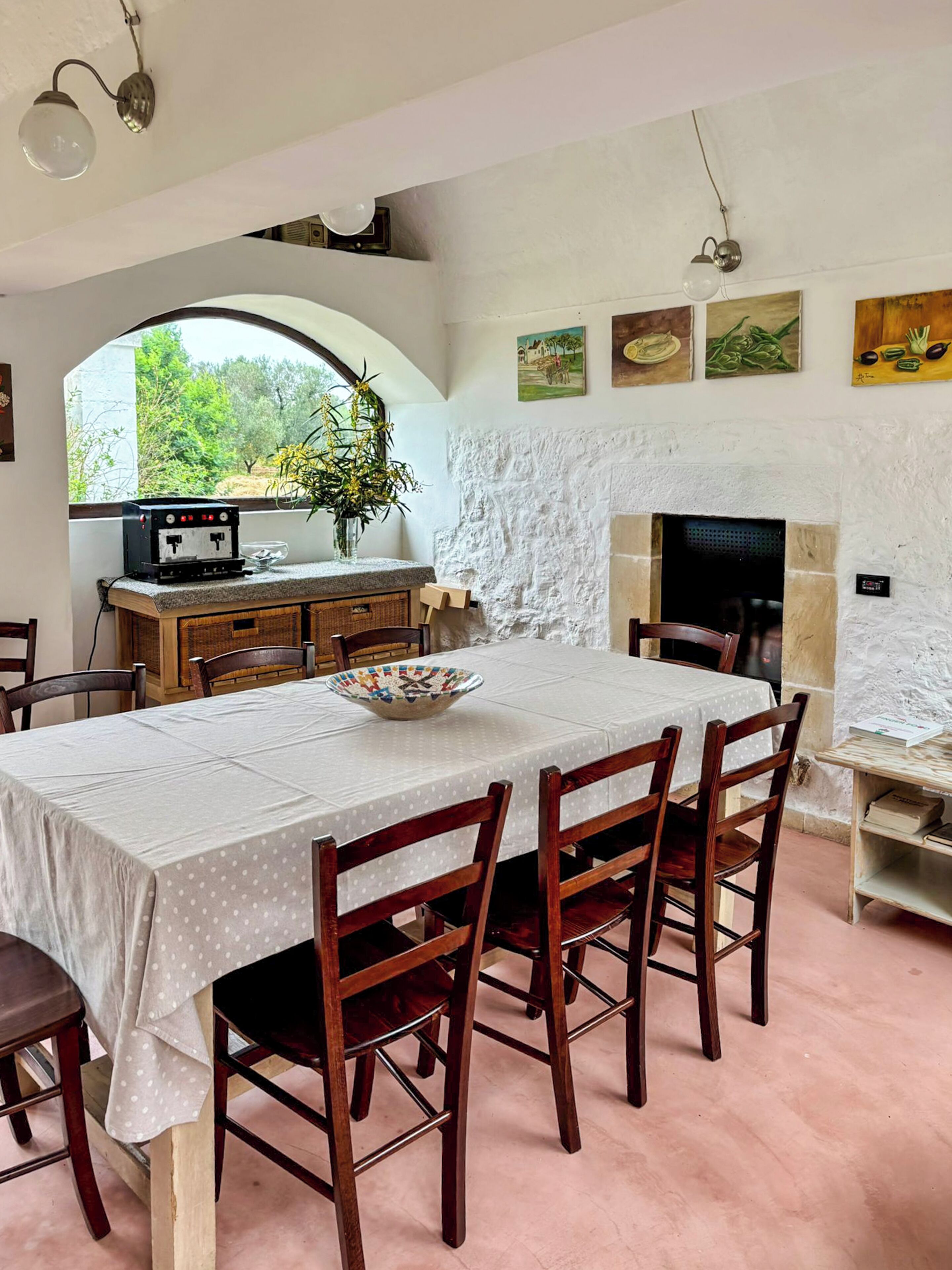 Uma sala de jantar com uma mesa posta para seis pessoas, cadeiras de madeira, toalha de mesa branca, um vaso com flores, uma área de cozinha com fogão e uma lareira.