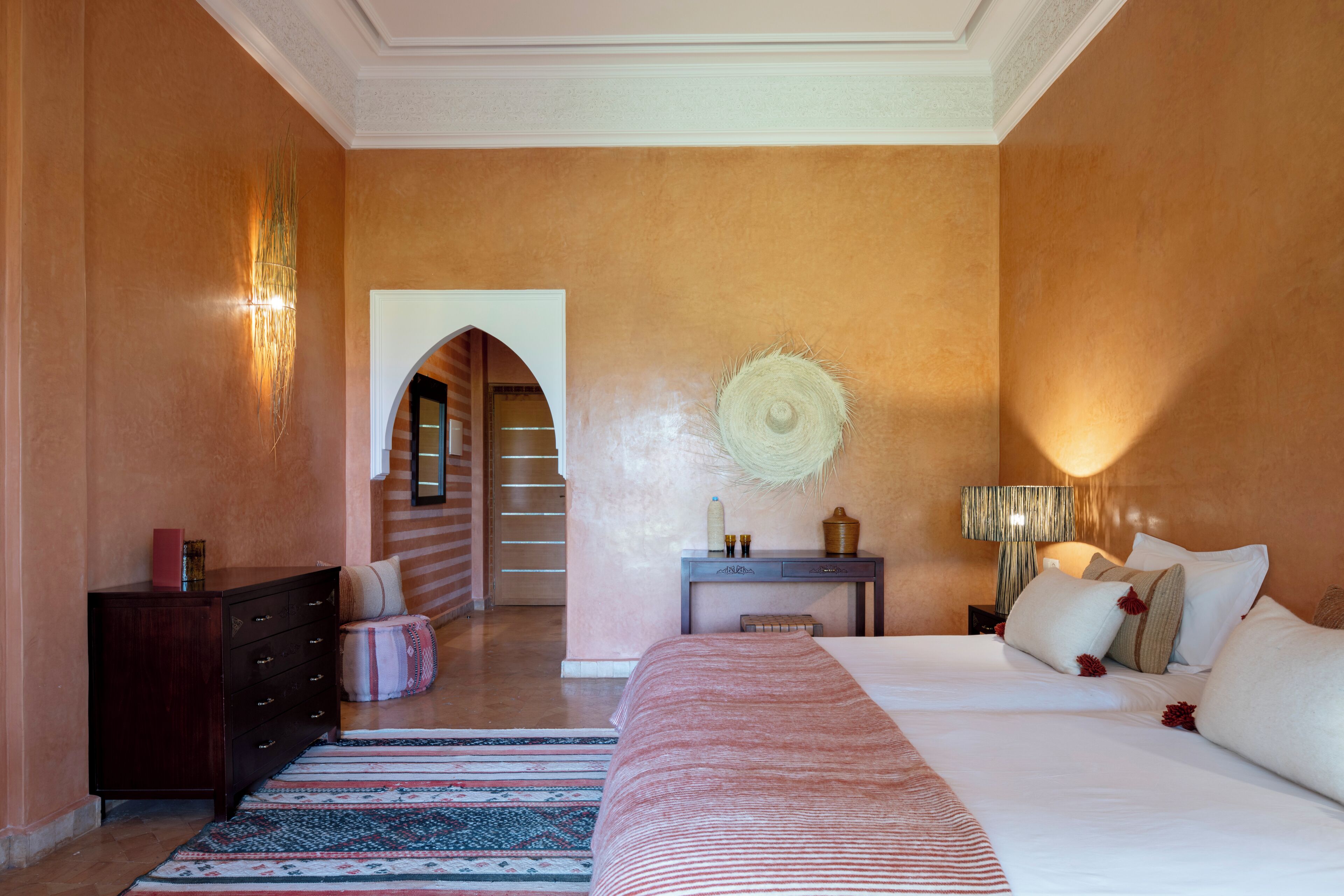 Quarto com cama, mesa de cabeceira, abajur, cômoda, tapete e uma porta em arco.