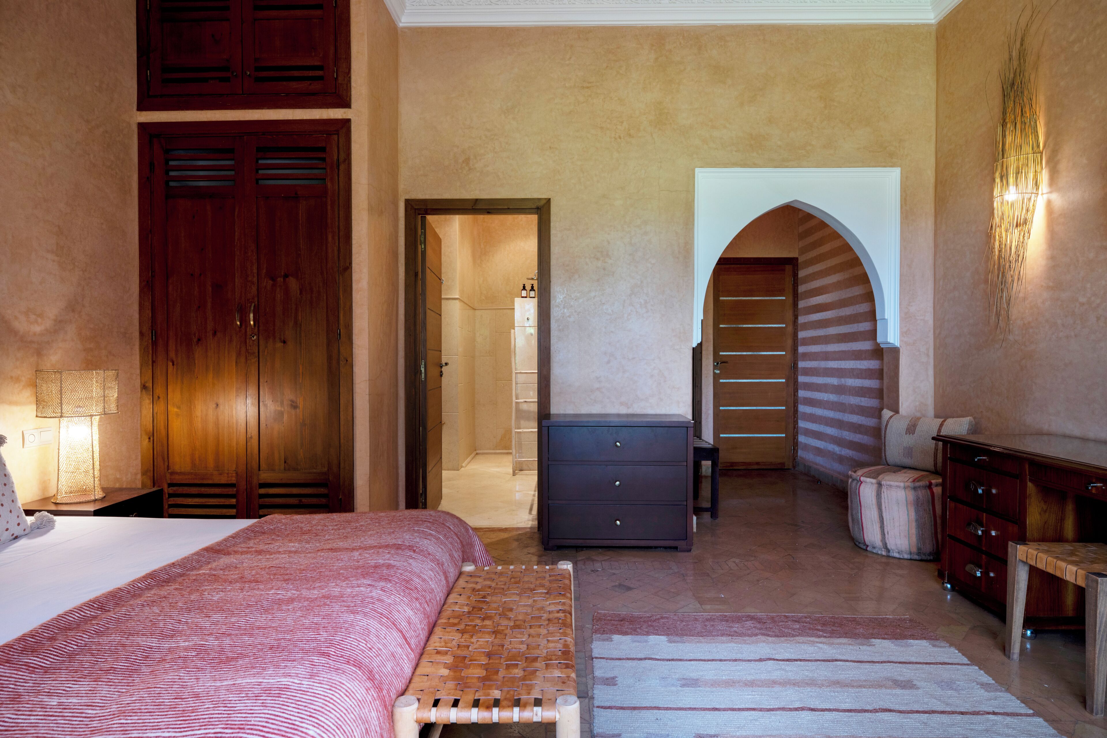 Um quarto com cama, guarda-roupa de madeira, um banco, penteadeira e uma porta em arco que dá para o banheiro.