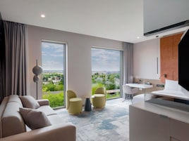 Quarto de hotel moderno com uma janela ampla, um sofá, duas cadeiras, uma mesinha, uma escrivaninha e uma cama.