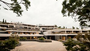 Um edifício moderno de vários andares com um pátio ajardinado, apresentando uma combinação de telhados planos e inclinados, e diversas plantas e árvores.
